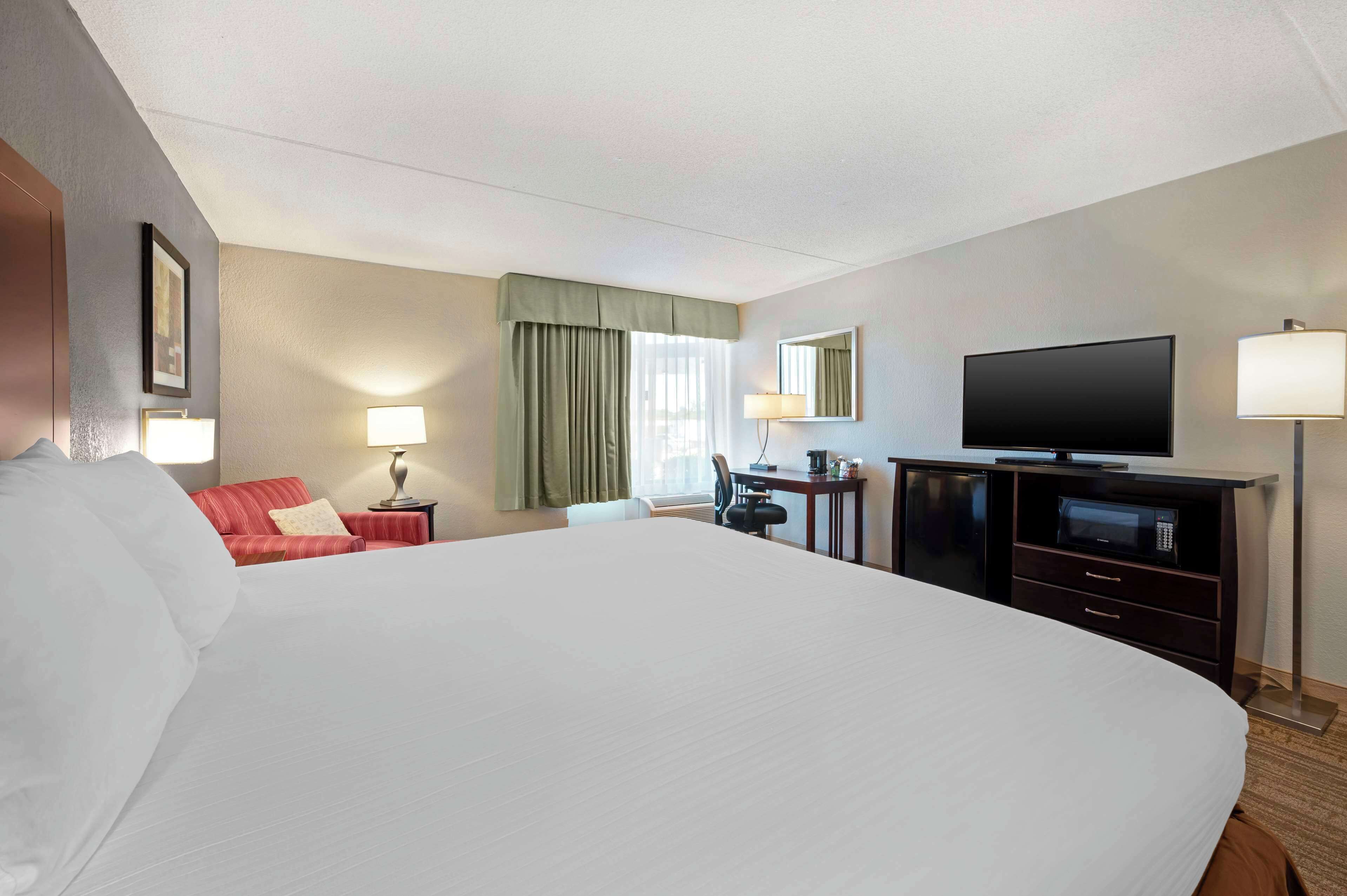 Quarto de hotel com uma cama grande, uma escrivaninha com televisão de tela plana, uma cadeira, um abajur, um sofá vermelho e uma janela com cortinas.
