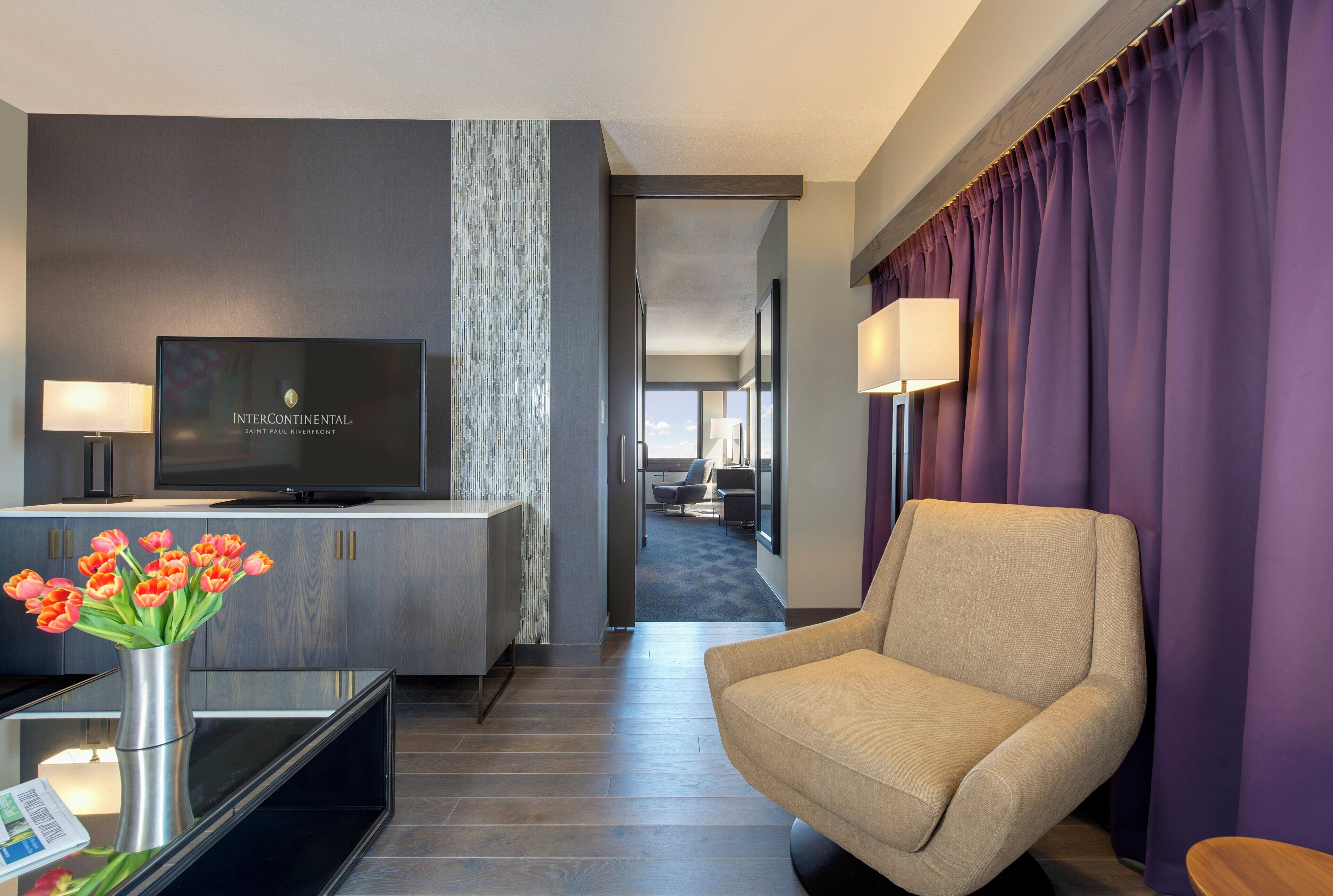 Quarto de hotel com televisão de tela plana, escrivaninha com abajur, cadeira e vista para o corredor.