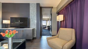 Quarto de hotel com televisão de tela plana, escrivaninha com abajur, cadeira e vista para o corredor.
