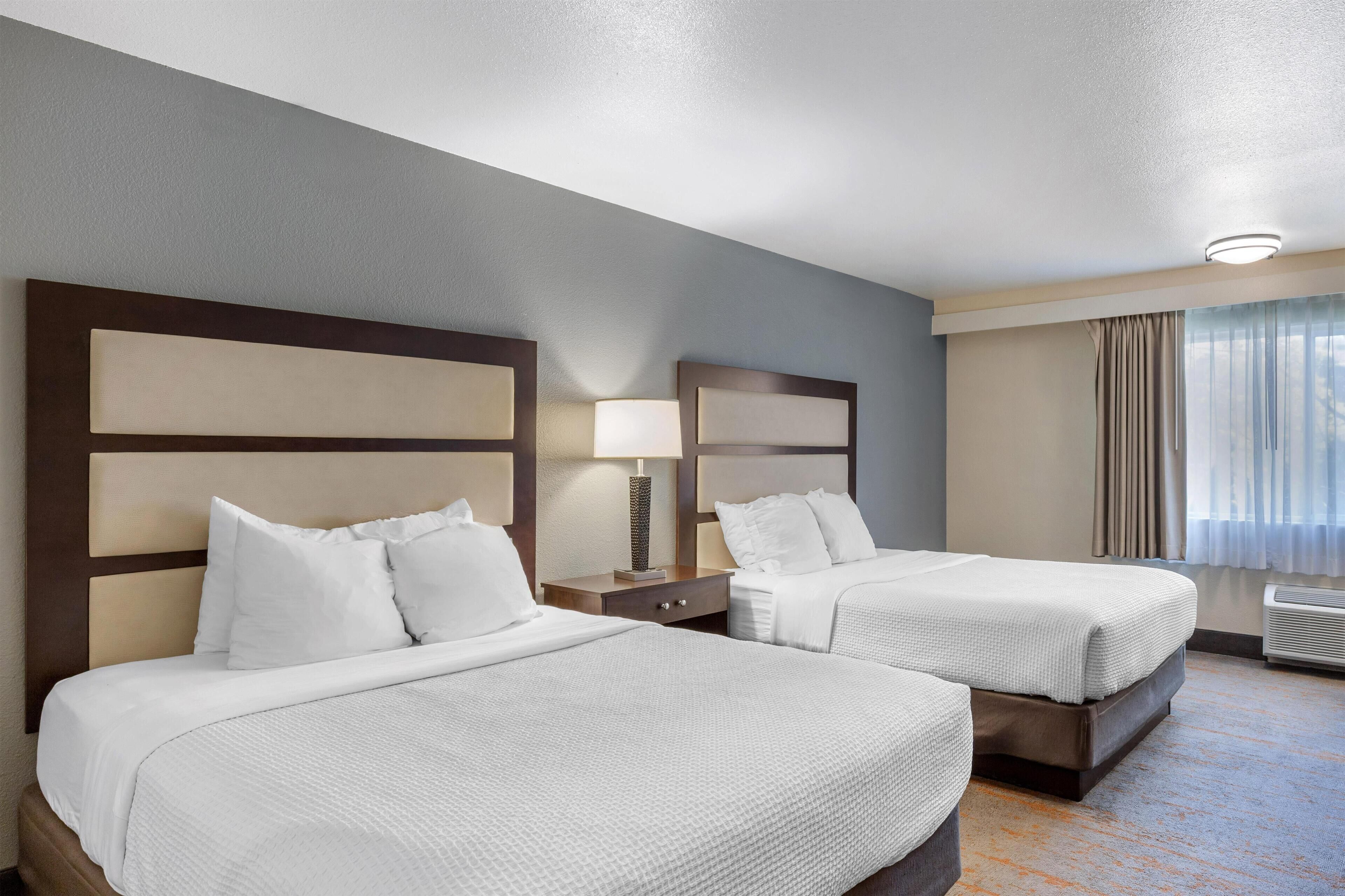 Quarto de hotel com duas camas, uma mesa de cabeceira com abajur, janela com cortinas e luminária no teto.