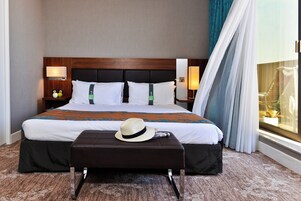 Quarto de hotel com uma cama grande, duas luminárias de cabeceira, uma mesa de cabeceira, uma janela com cortinas e uma pequena mesa com um chapéu.