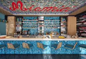 Um balcão com mosaico de azulejos azuis, prateleiras cheias de garrafas e banquetas de vime.