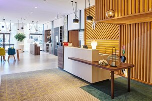 Uma área de recepção de hotel com balcão de madeira, bancada de atendimento e área de espera com cadeiras.