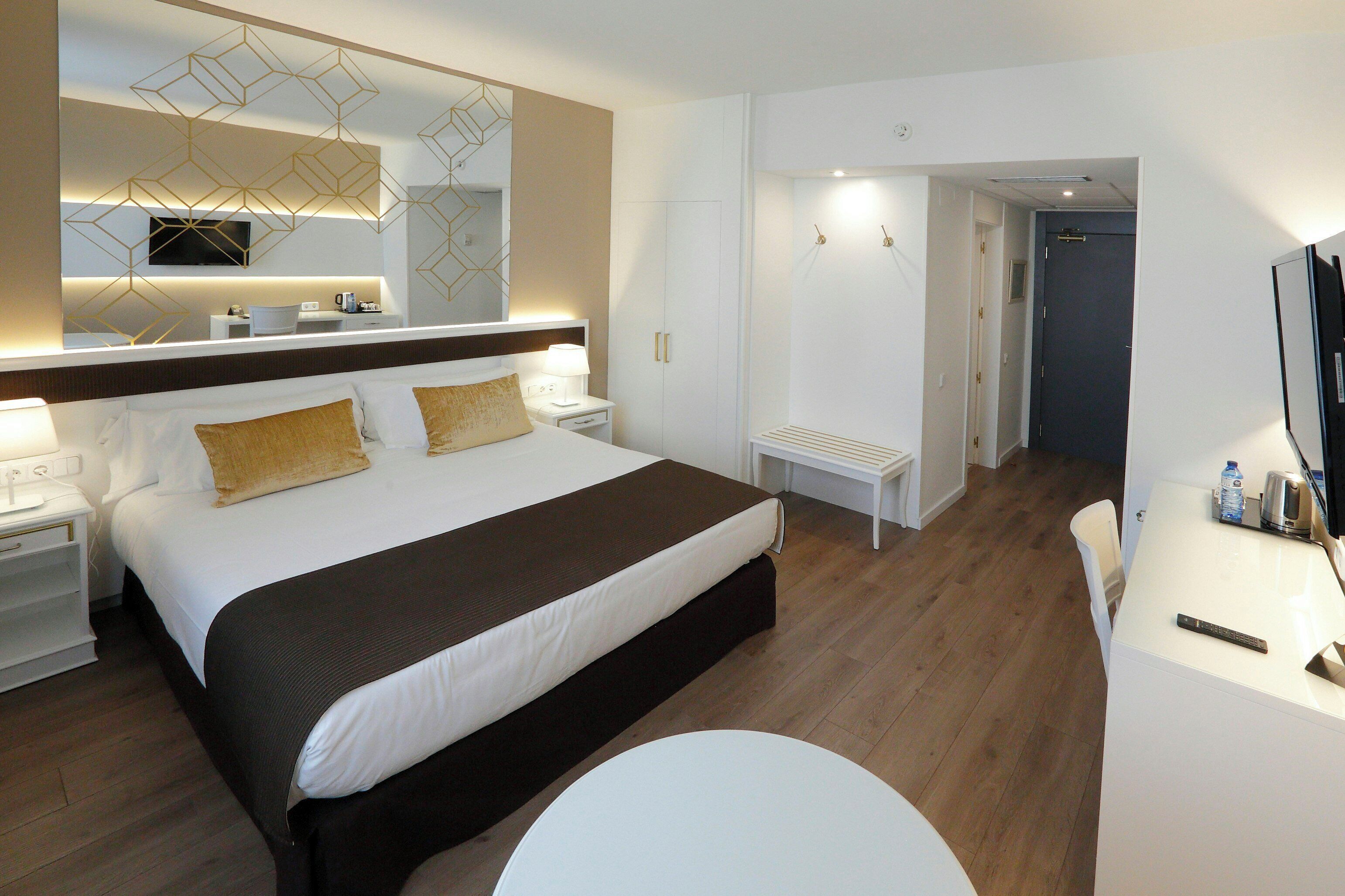 Quarto de hotel com cama, mesas de cabeceira, escrivaninha, cadeira, mesa redonda, espelho e porta.