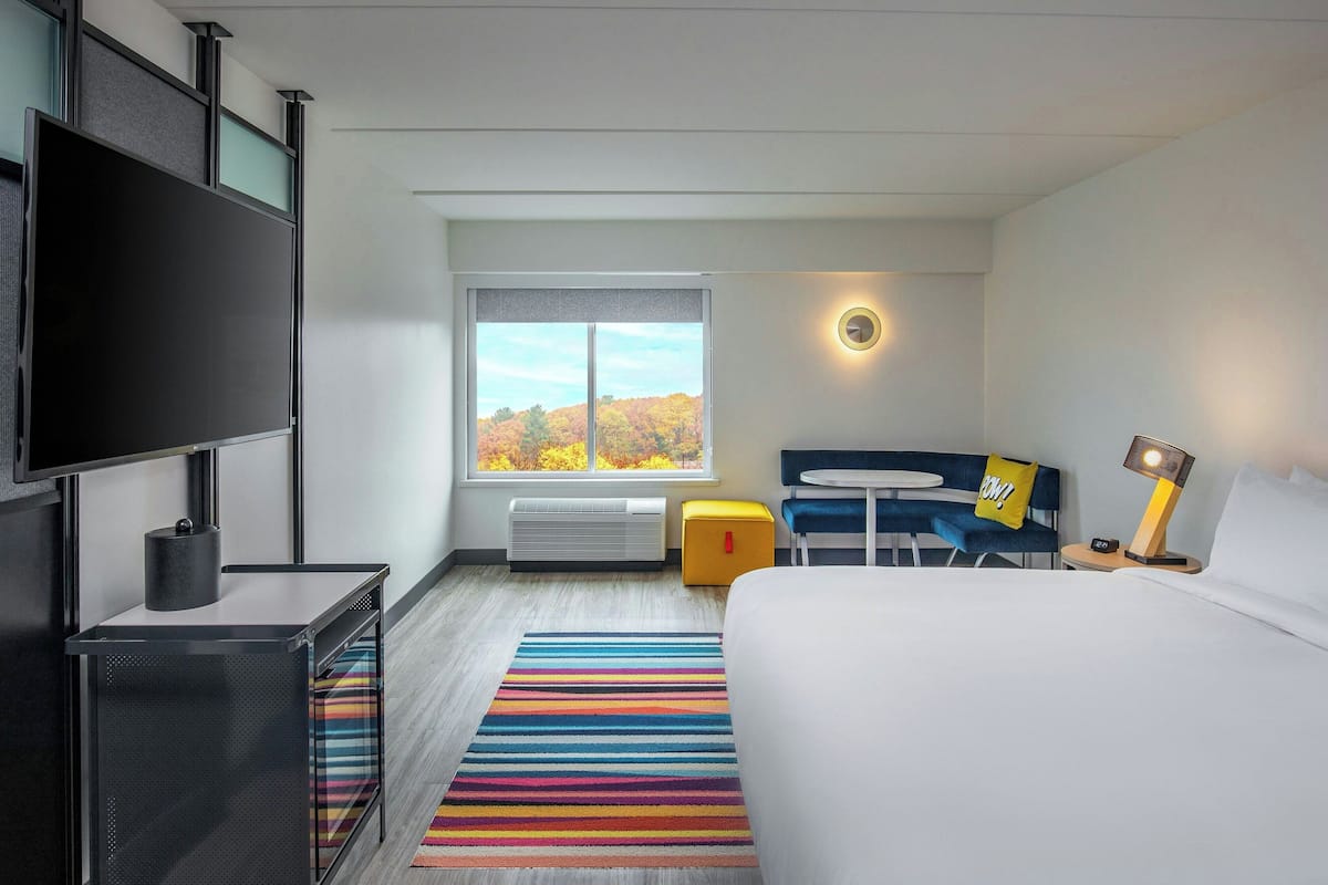Quarto de hotel com cama, televisão, escrivaninha, cadeira, mesinha e janela com vista para árvores de outono.