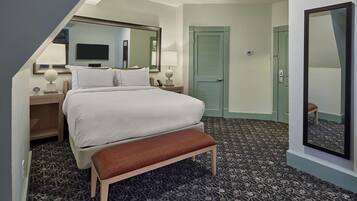 Quarto de hotel com uma cama grande, um banco, uma escrivaninha com uma luminária, um espelho e uma porta.