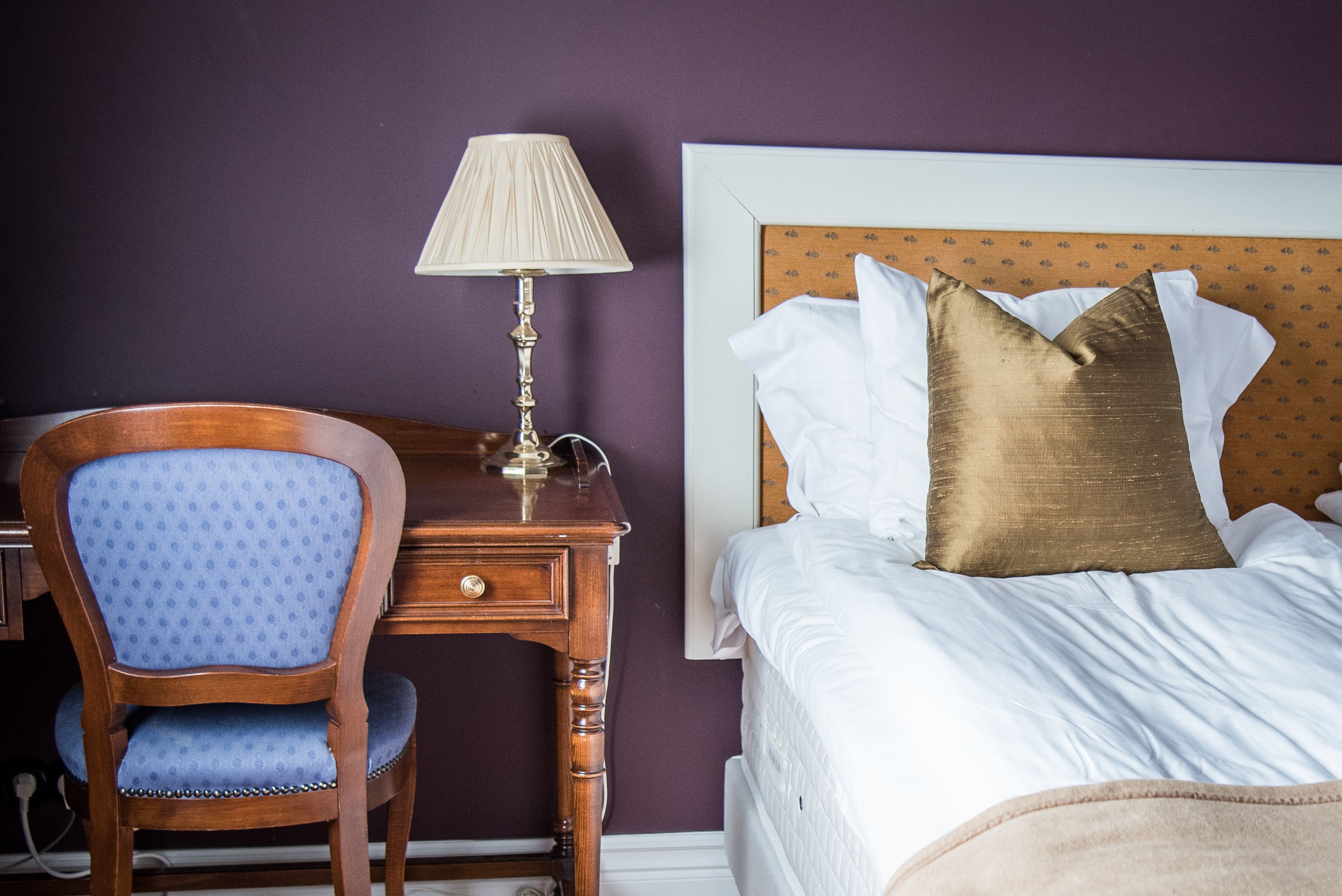 Quarto de hotel com cama, mesa de cabeceira de madeira, luminária e poltrona azul estofada.