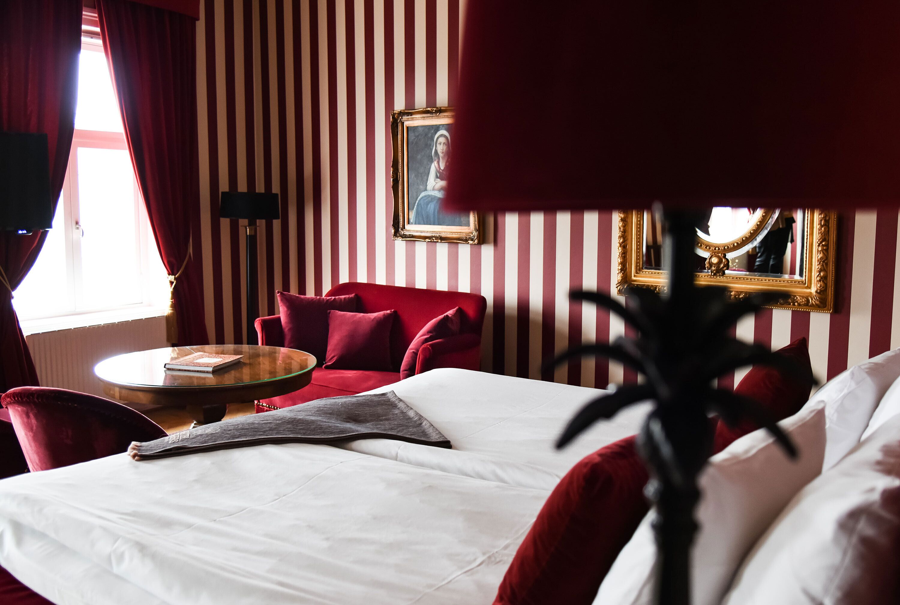 Quarto de hotel com uma cama grande, cadeiras estofadas em vermelho, uma mesa redonda e um espelho com moldura dourada.