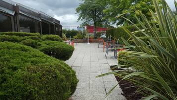 Um espaço ao ar livre pavimentado com sebes bem aparadas, mobiliário de jardim com cadeiras laranja e cinzas, e um edifício com fachada de vidro ao fundo.