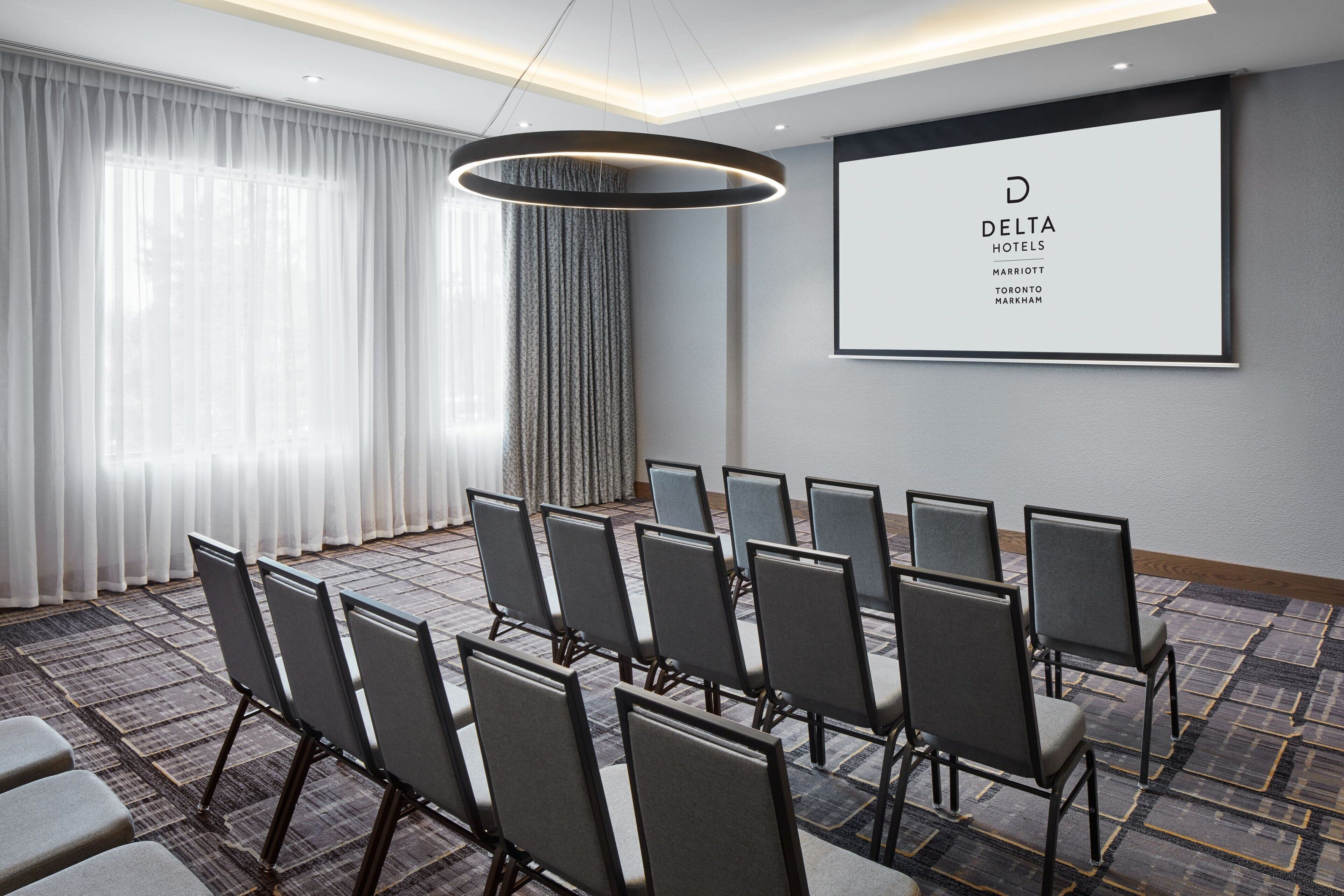 Uma sala de conferências com fileiras de cadeiras, uma tela grande exibindo 'Delta Hotels Marriott Toronto Markham' e um moderno candeeiro de teto.