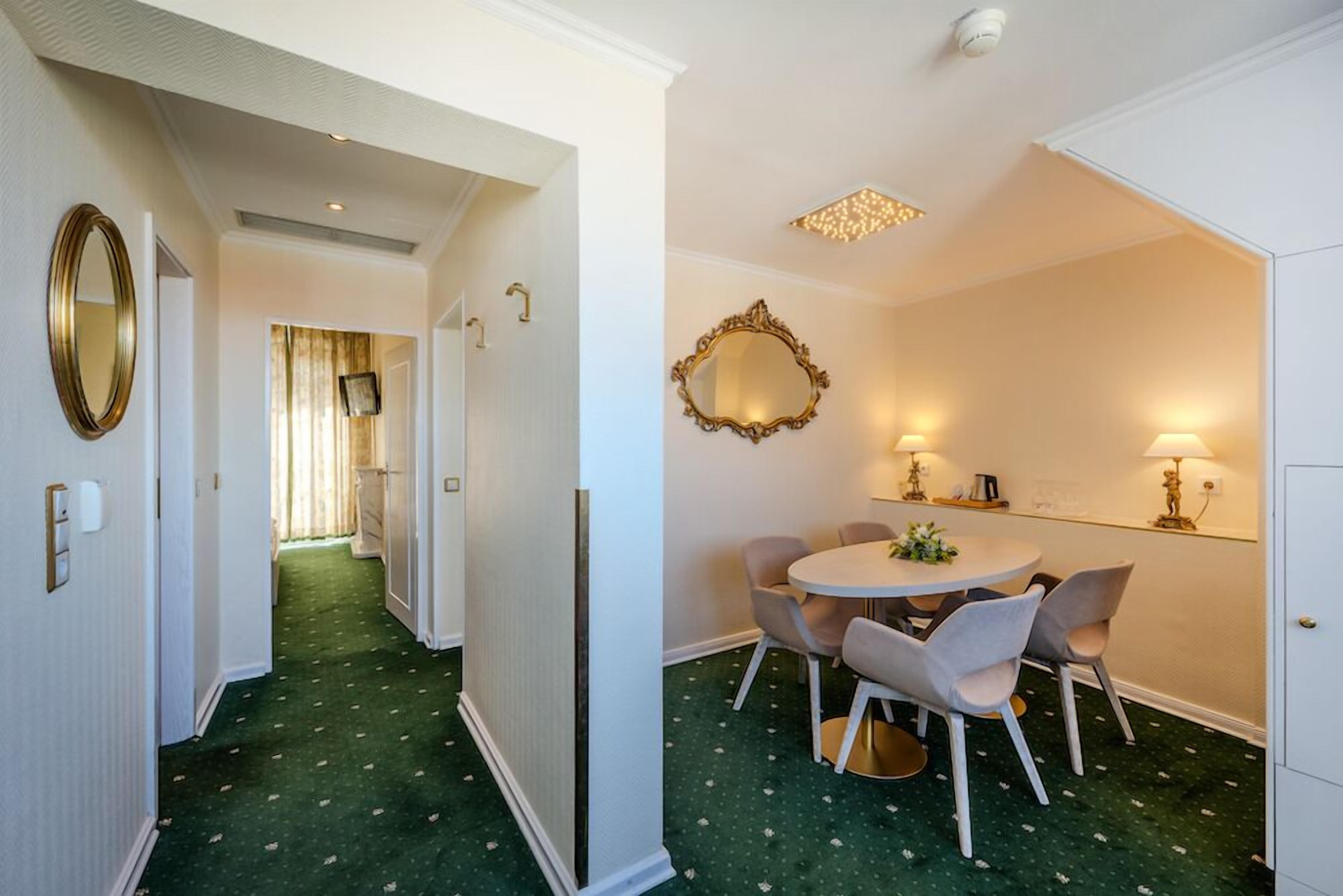 Quarto de hotel com carpete verde, mesa de jantar redonda com cadeiras, um espelho grande e um candelabro.