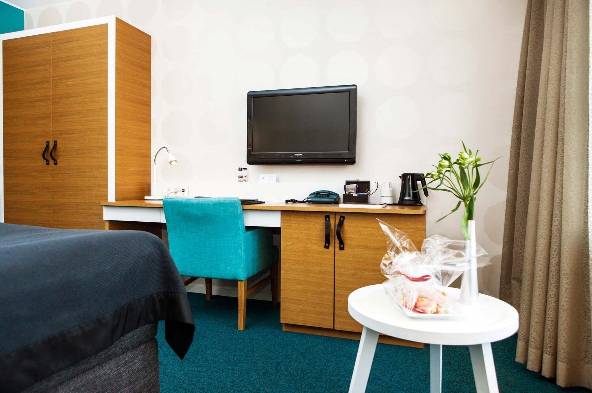 Quarto de hotel com uma cama, uma escrivaninha com um computador, uma televisão, uma cafeteira e uma mesinha com uma planta e objetos embalados.