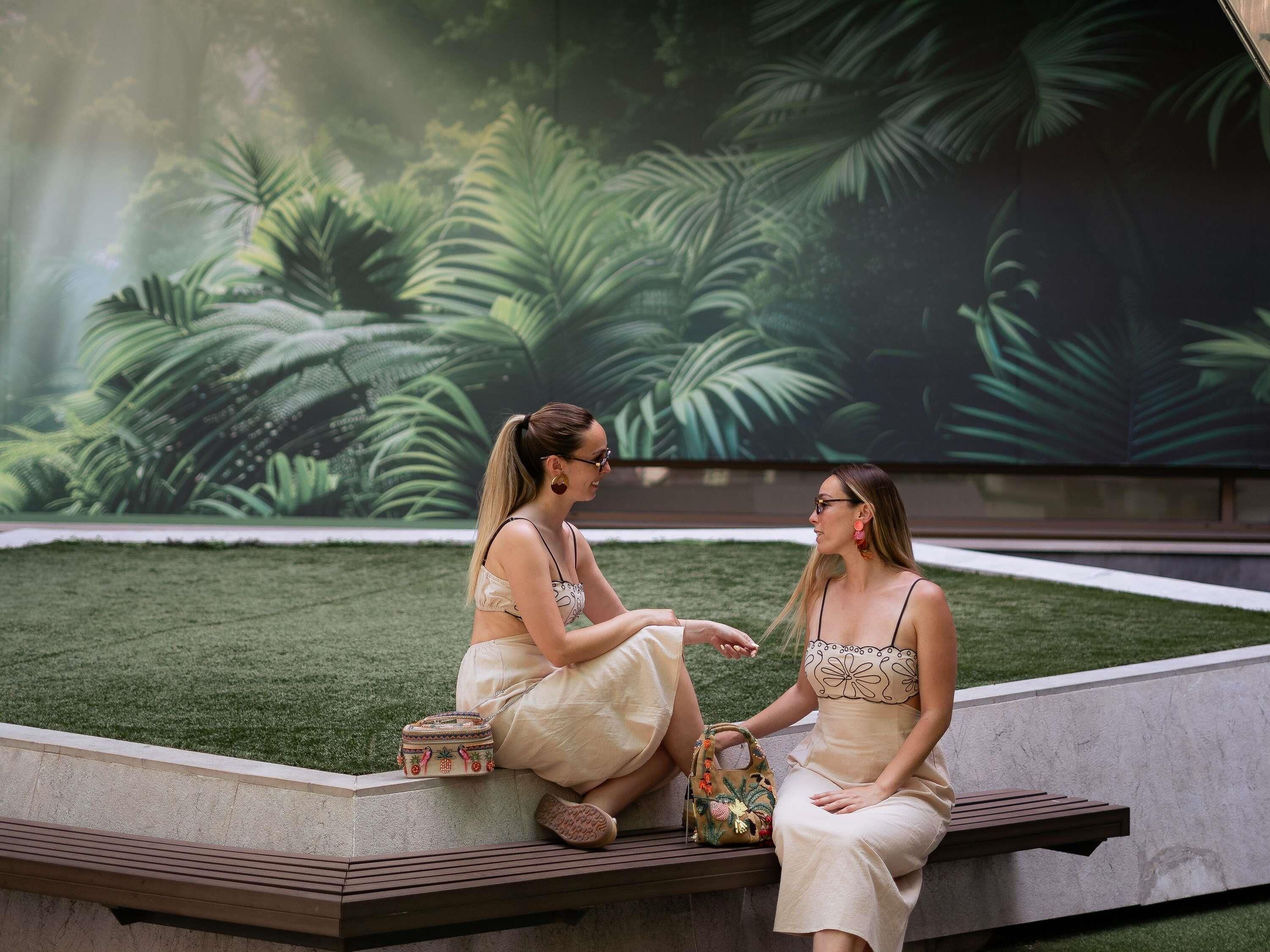 Duas pessoas sentadas num banco ao ar livre, com folhagem verde ao fundo.