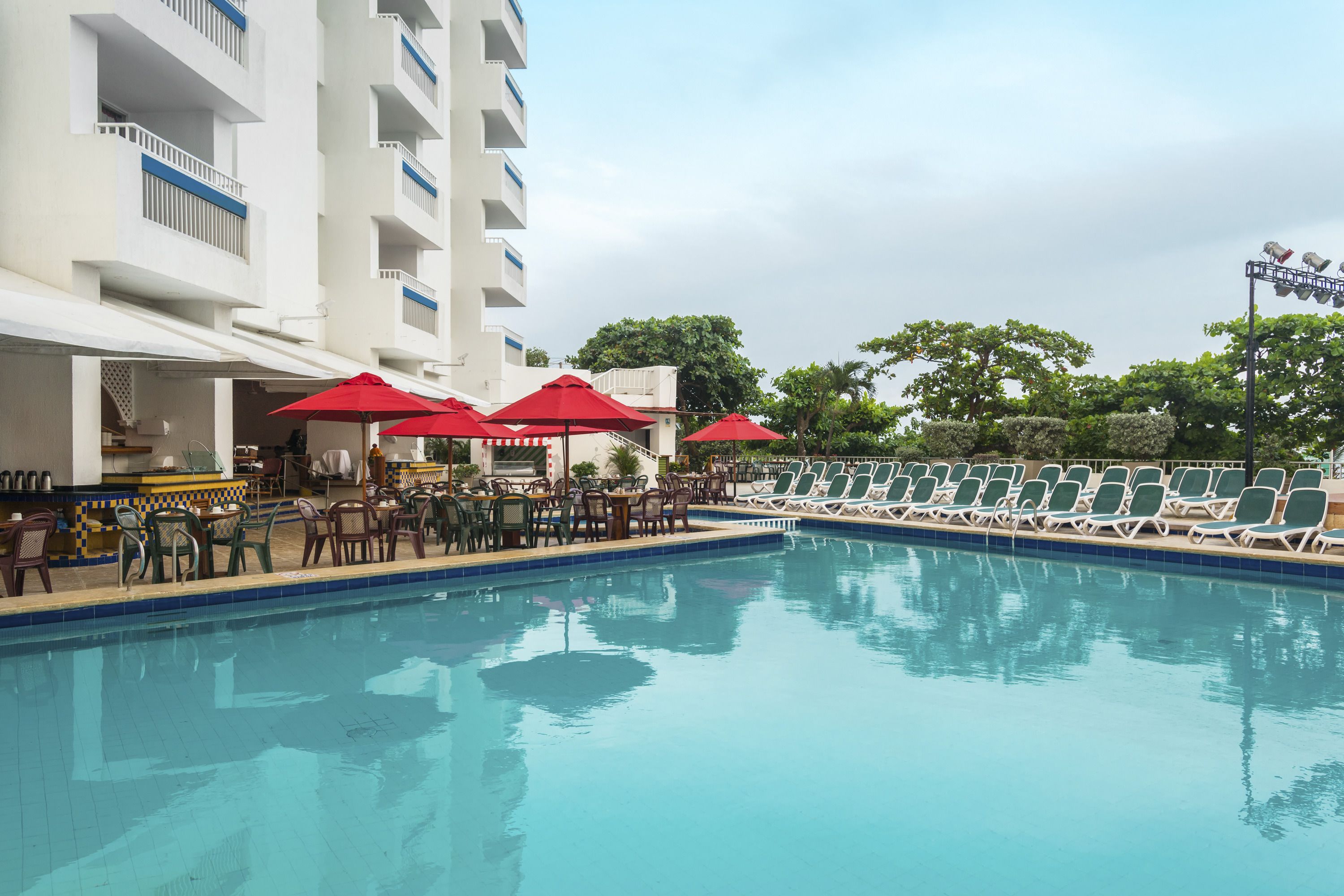 Piscina com espreguiçadeiras e guarda-sóis vermelhos, rodeada por um edifício com várias andares e varandas.