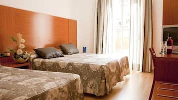 Quarto de hotel com duas camas, cabeceira de madeira, escrivaninha com uma garrafa e copos, e janela com cortinas.