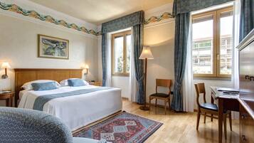 Quarto de hotel com uma cama grande, uma escrivaninha, uma cadeira, um abajur e vista para o exterior.