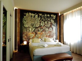 Quarto de hotel com uma cama grande, um banco, uma mesa de cabeceira com abajur e uma parede decorativa.