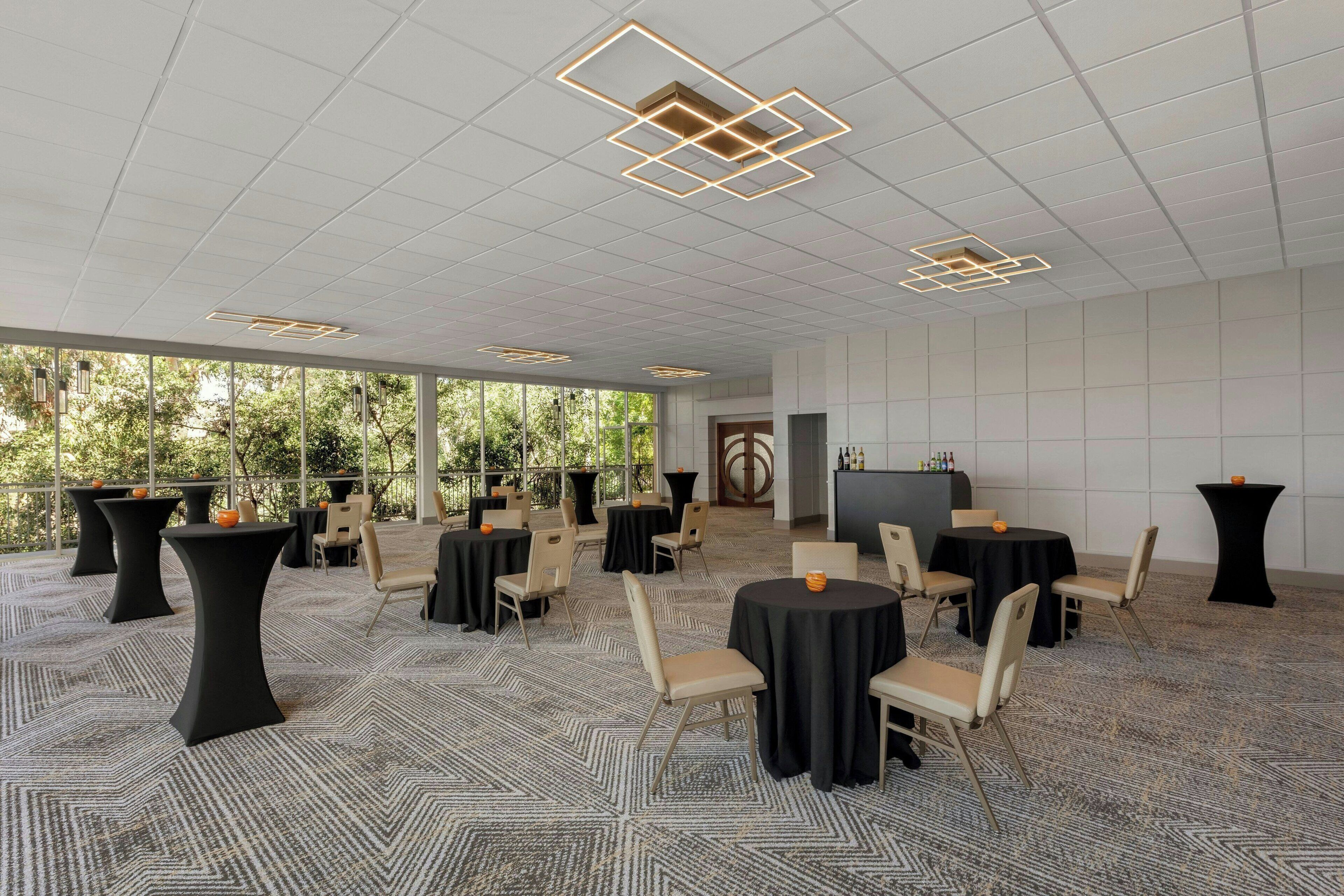 Um espaço amplo e interno com várias mesas redondas e cadeiras, um tapete estampado e luminárias geométricas no teto.