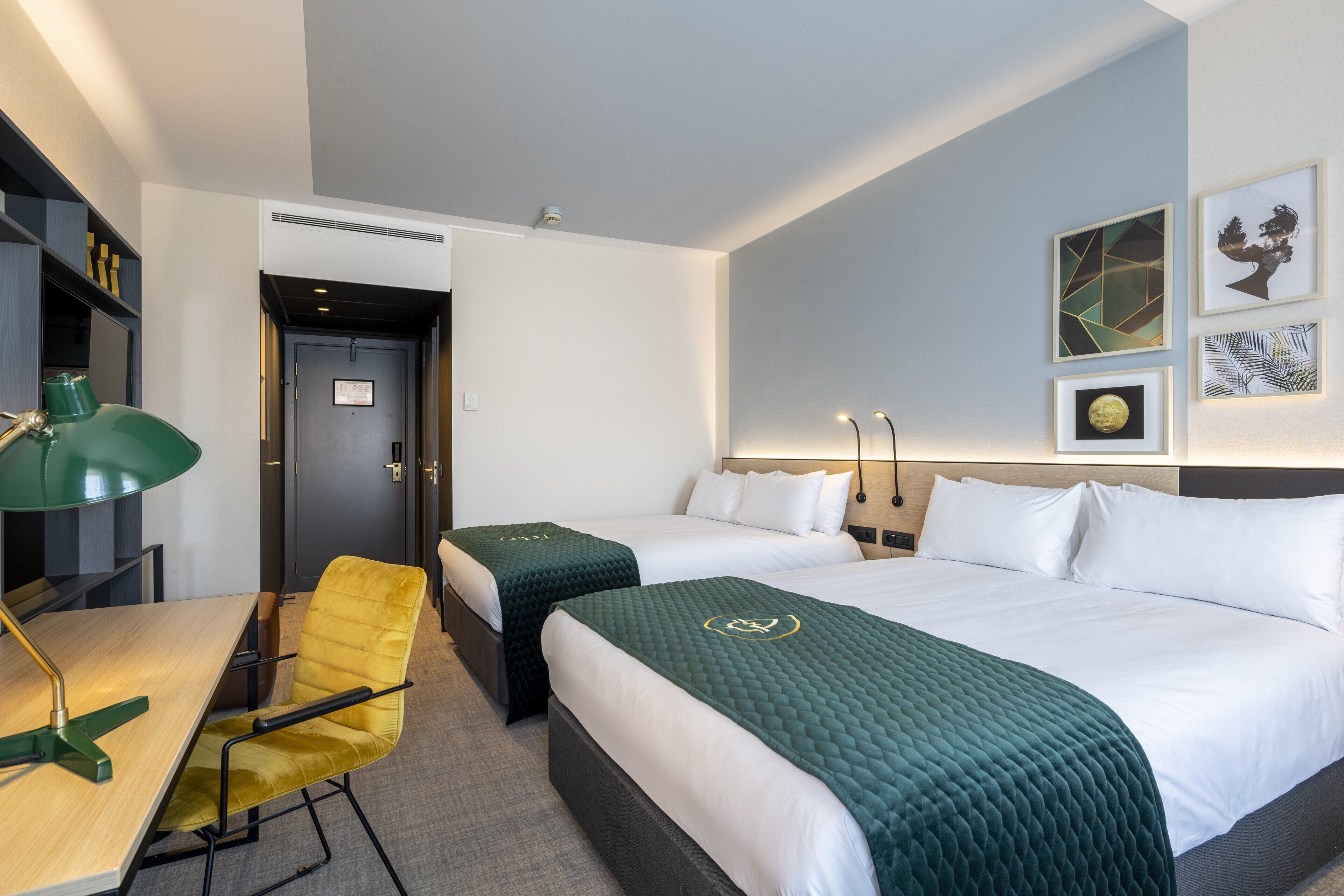 Quarto de hotel com duas camas, uma escrivaninha, uma cadeira e uma luminária de mesa. Há uma porta que dá para outro quarto.