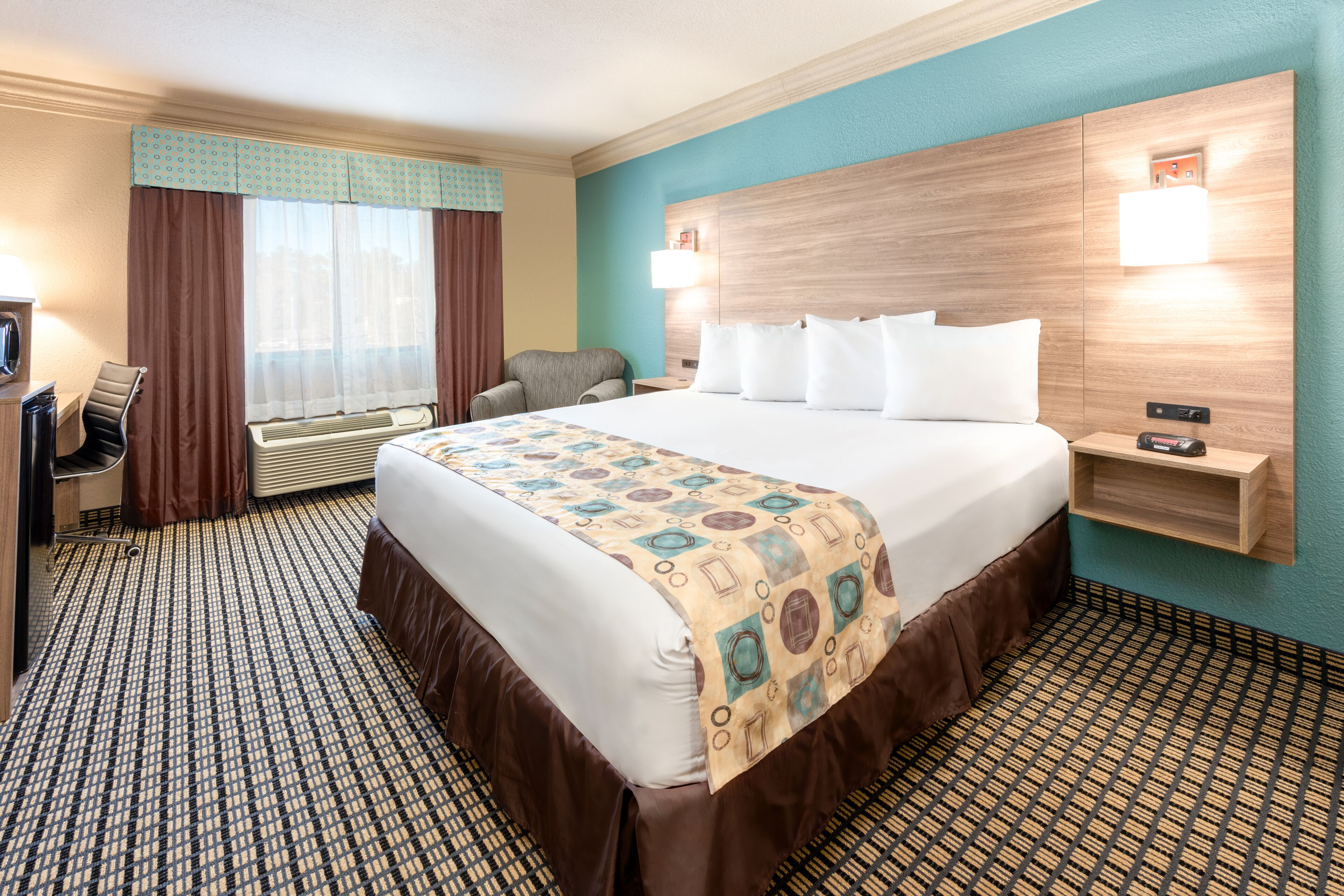Quarto de hotel com uma cama grande, uma secretária com cadeira, uma janela com cortinas e uma estante fixada na parede.