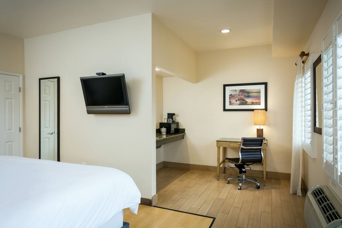 Quarto de hotel com uma cama, uma escrivaninha com cadeira, uma televisão, um quadro na parede e uma janela com persianas.