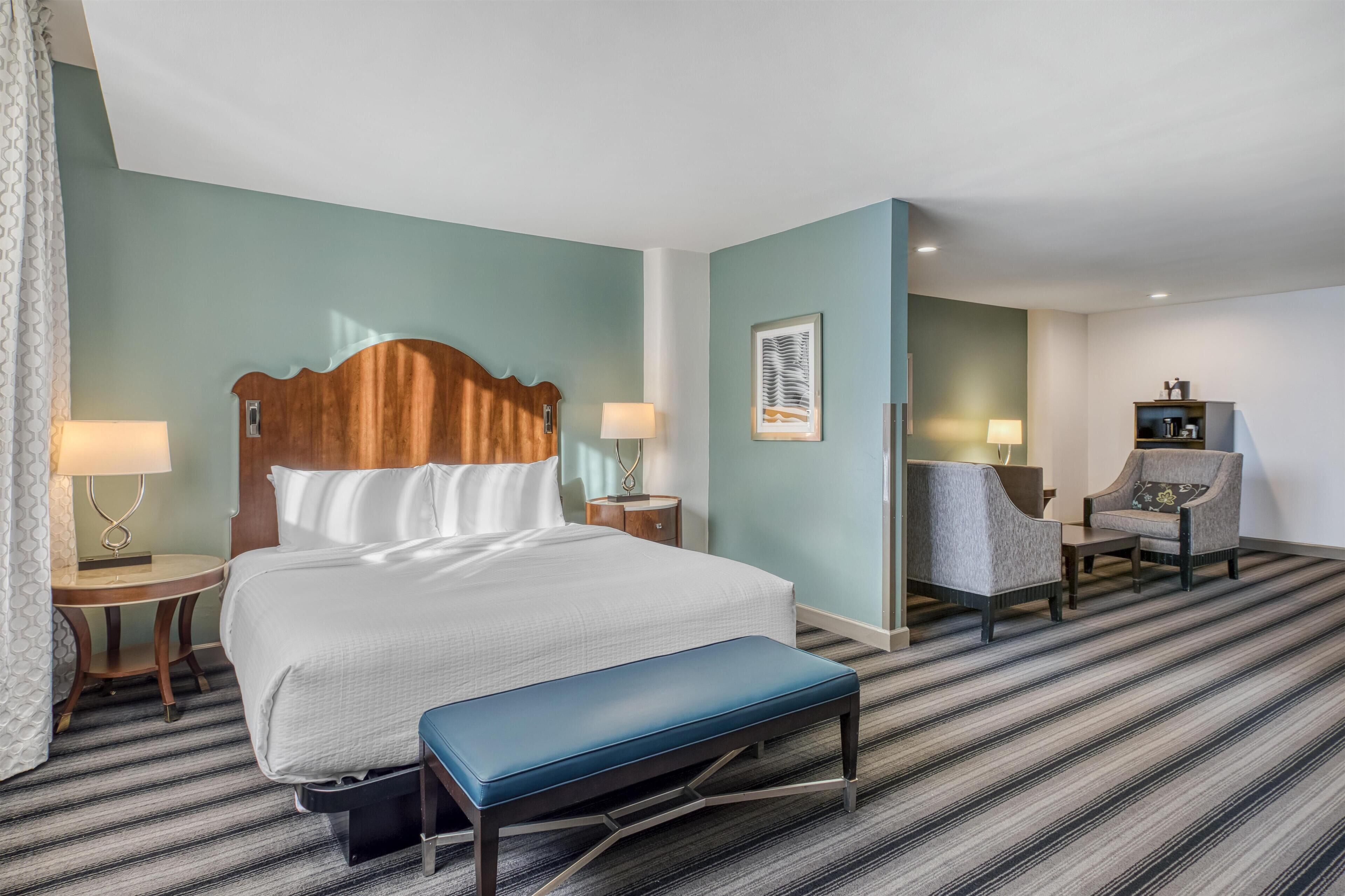 Quarto de hotel com uma cama grande, mesas de cabeceira, um banco, uma cadeira e uma pequena mesa com abajur.
