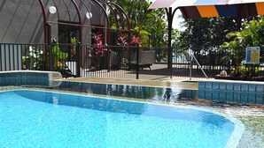 Piscina com água azul cristalina, cercada por uma grade e uma área coberta com um banco.