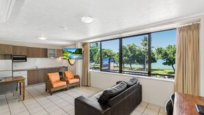 Quarto de hotel moderno com cozinha, sofá, mesa de jantar e uma grande janela com vista para uma paisagem tropical.