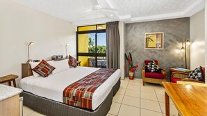 Quarto de hotel com uma cama grande, uma escrivaninha, uma cadeira, uma mesinha, um ventilador de teto, uma janela com cortinas e um aplique na parede.