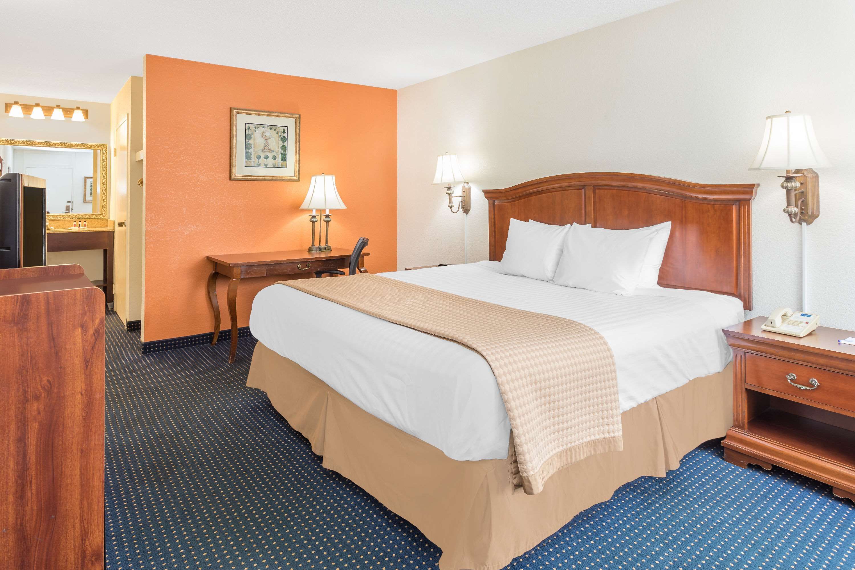 Quarto de hotel com uma cama grande, uma escrivaninha, uma cadeira, uma luminária de mesa, uma mesinha de cabeceira com abajur e um telefone.