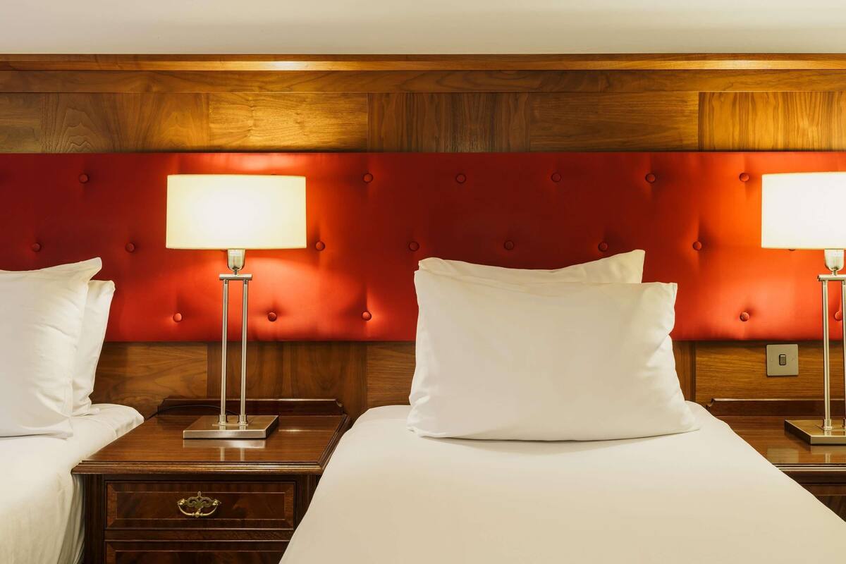 Quarto de hotel com duas camas, cada uma acompanhada de uma mesa de cabeceira de madeira, uma luminária e uma gaveta.