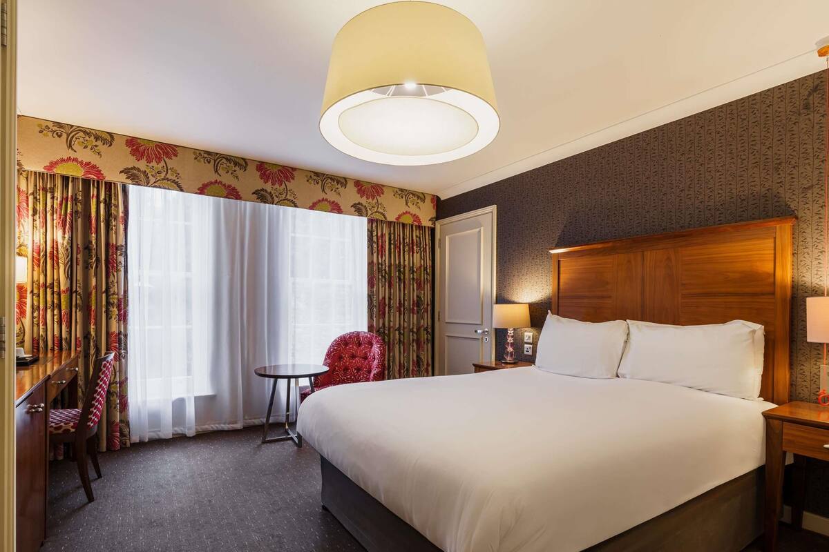 Quarto de hotel com uma cama grande, uma escrivaninha com cadeira, uma mesinha e uma porta.