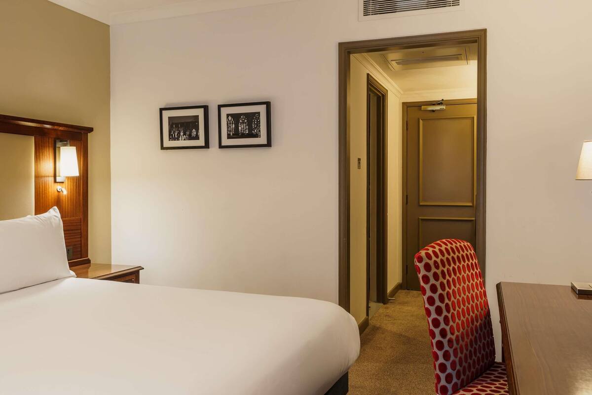 Quarto de hotel com cama, mesa de cabeceira, uma cadeira e uma mesa. Há duas fotos emolduradas na parede.