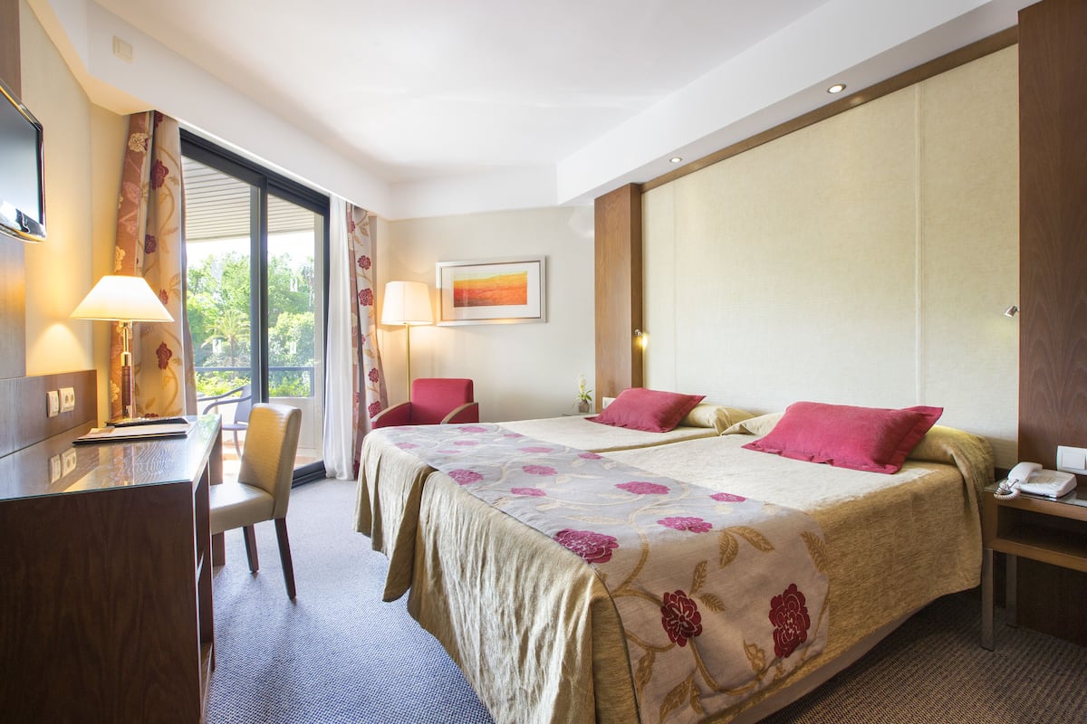 Quarto de hotel com duas camas, uma escrivaninha, uma cadeira e uma janela com cortinas.