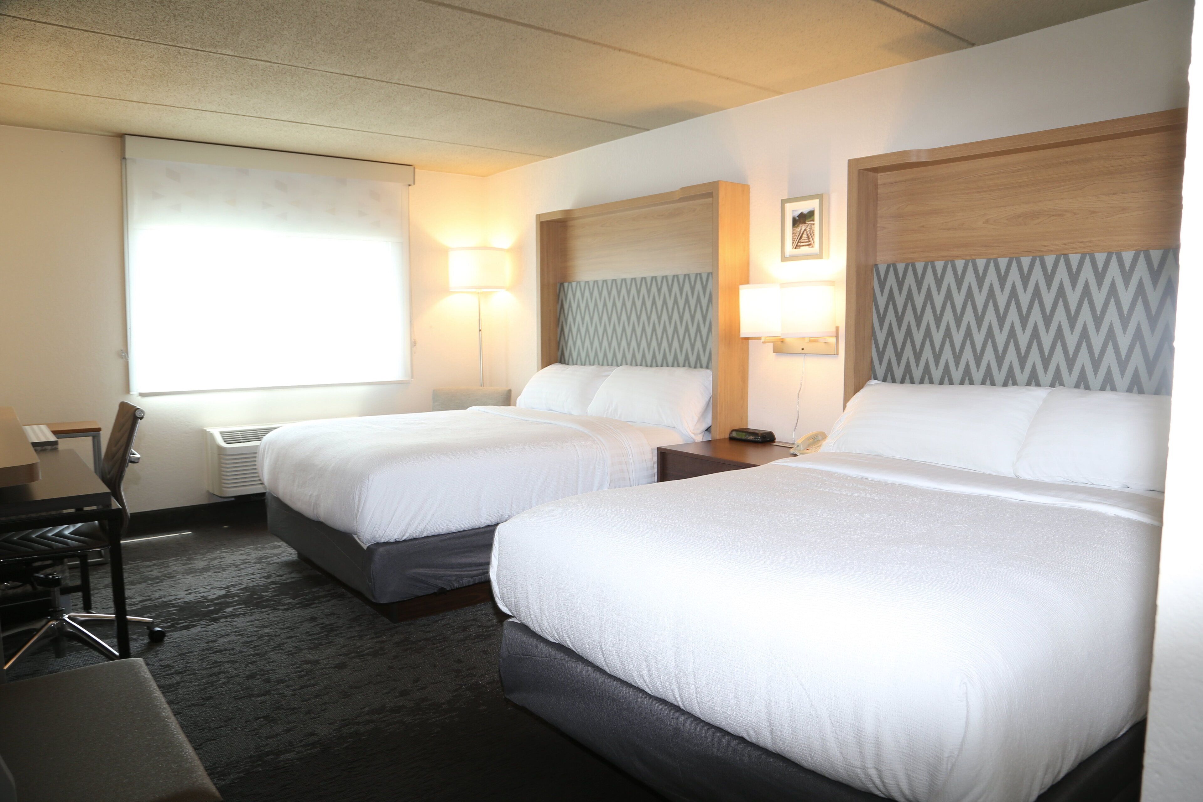 Quarto de hotel com duas camas, uma escrivaninha, uma cadeira e uma janela com persianas.