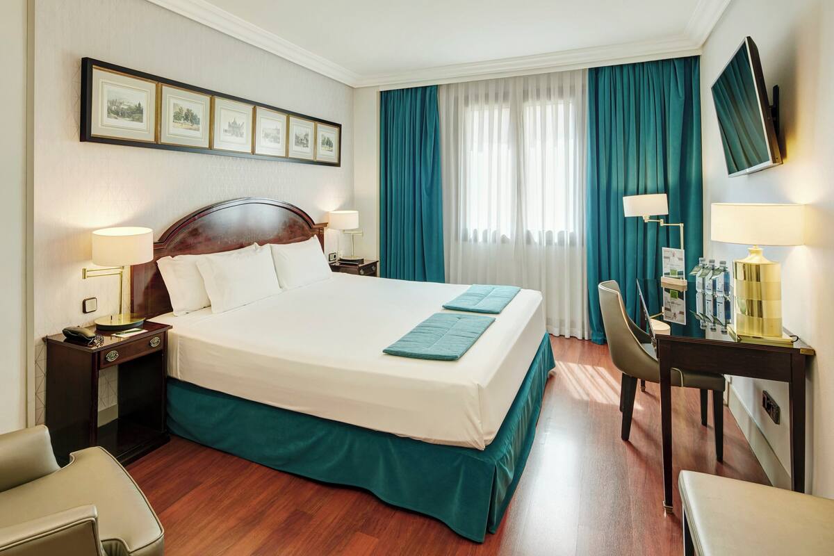 Quarto de hotel com uma cama grande, duas mesas de cabeceira com luminárias, uma escrivaninha com cadeira, uma TV fixada na parede e uma janela com cortinas.