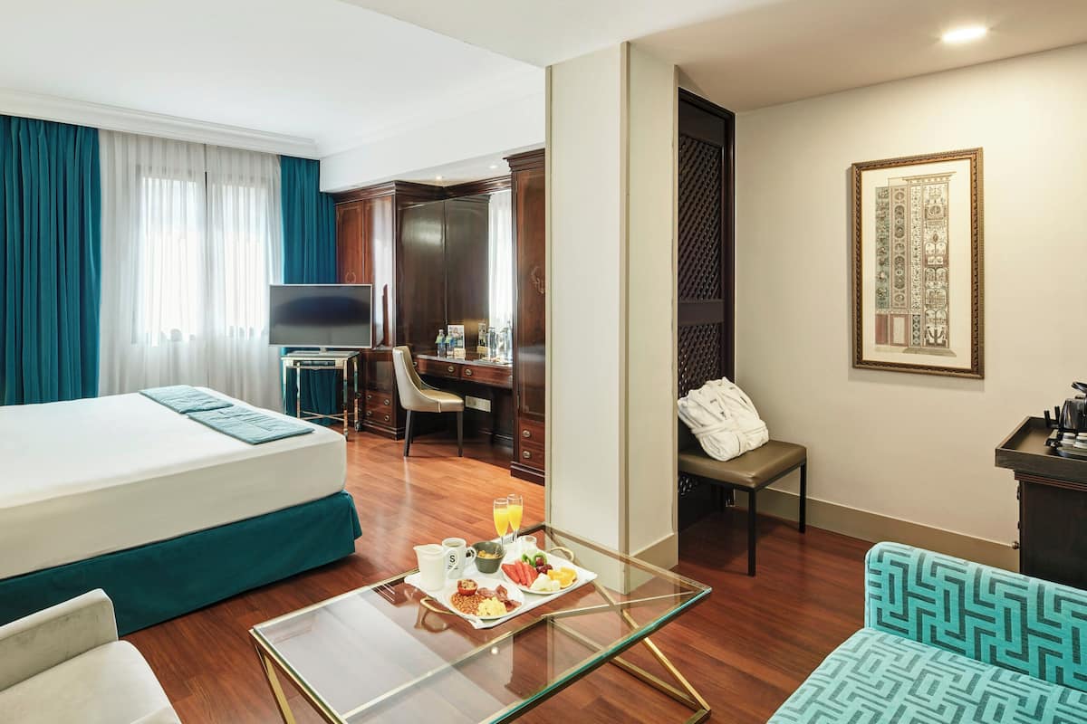Quarto de hotel com cama, mesa de trabalho, cadeira, mesinha de vidro com café da manhã e televisão.