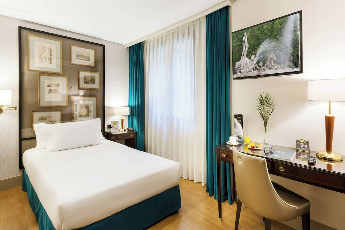 Quarto de hotel com cama, escrivaninha, cadeira, quadros emoldurados, cortinas, uma estátua e uma televisão.