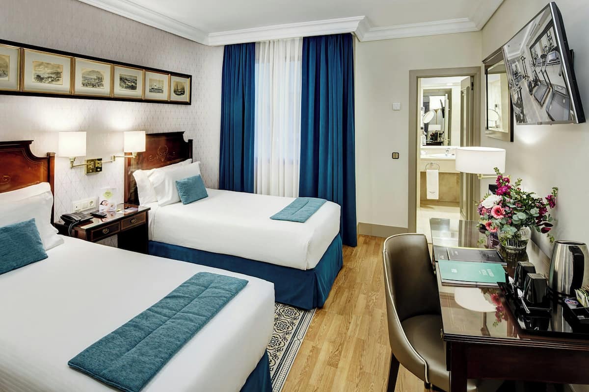 Quarto de hotel com duas camas, uma escrivaninha, uma cadeira, um espelho e uma parede com quadros.