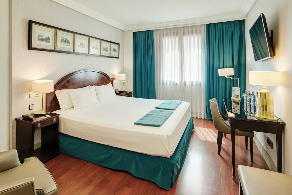 Quarto de hotel com uma cama grande, duas mesas de cabeceira com luminárias, uma escrivaninha com cadeira, uma TV fixada na parede e uma janela com cortinas.