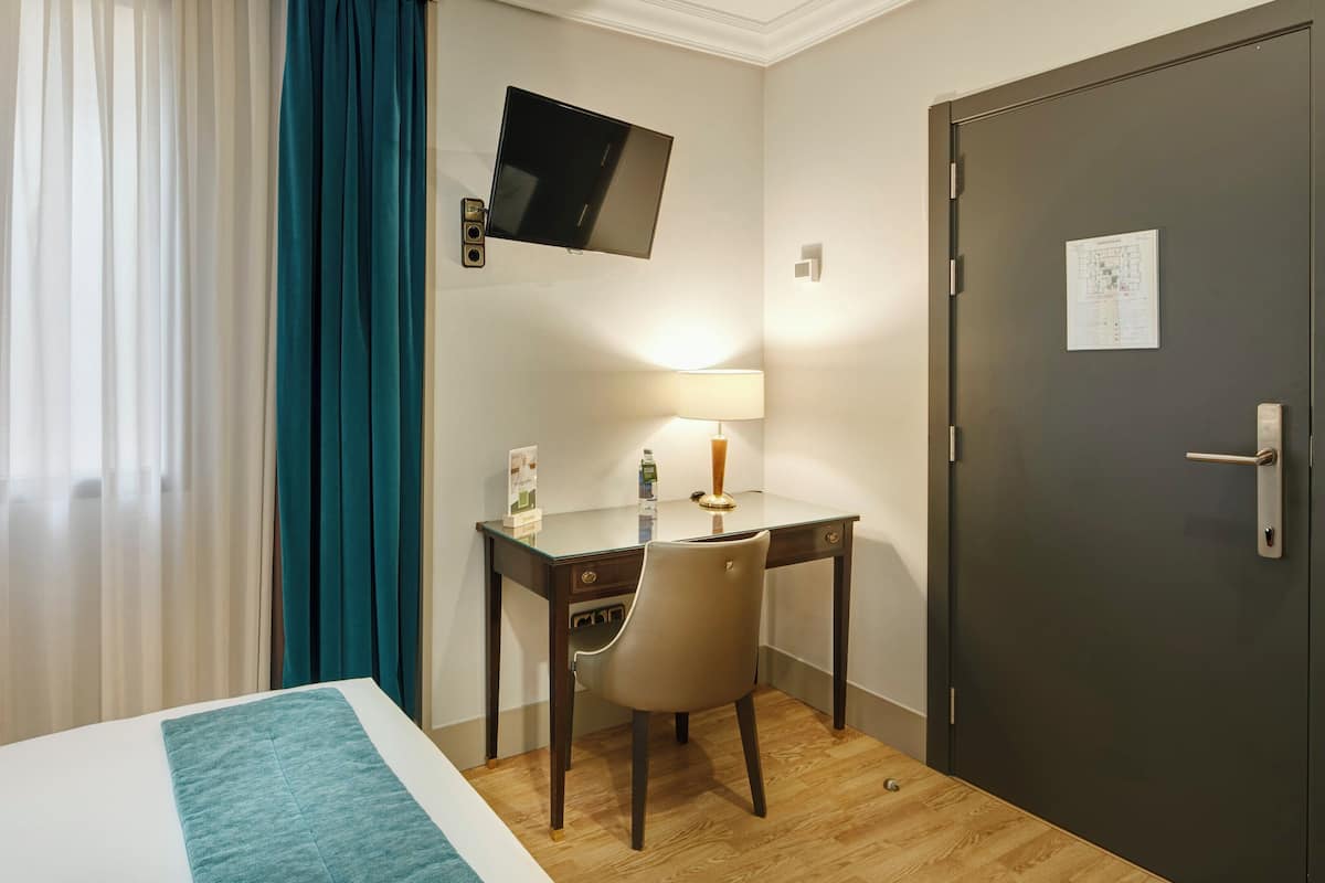 Quarto de hotel com cama, escrivaninha com cadeira, TV na parede e porta com leitor de cartão.