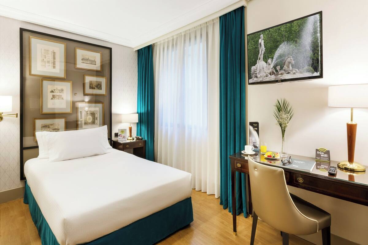Quarto de hotel com cama, escrivaninha, cadeira, quadros emoldurados, cortinas, uma estátua e uma televisão.