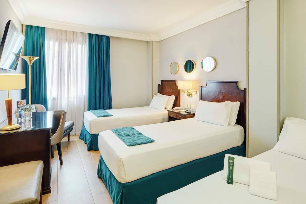 Quarto de hotel com duas camas, uma escrivaninha, uma cadeira, um abajur, um espelho e uma janela com cortinas.