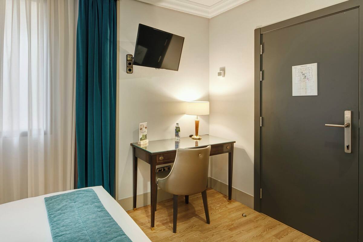 Quarto de hotel com cama, escrivaninha com cadeira, TV na parede e porta com leitor de cartão.