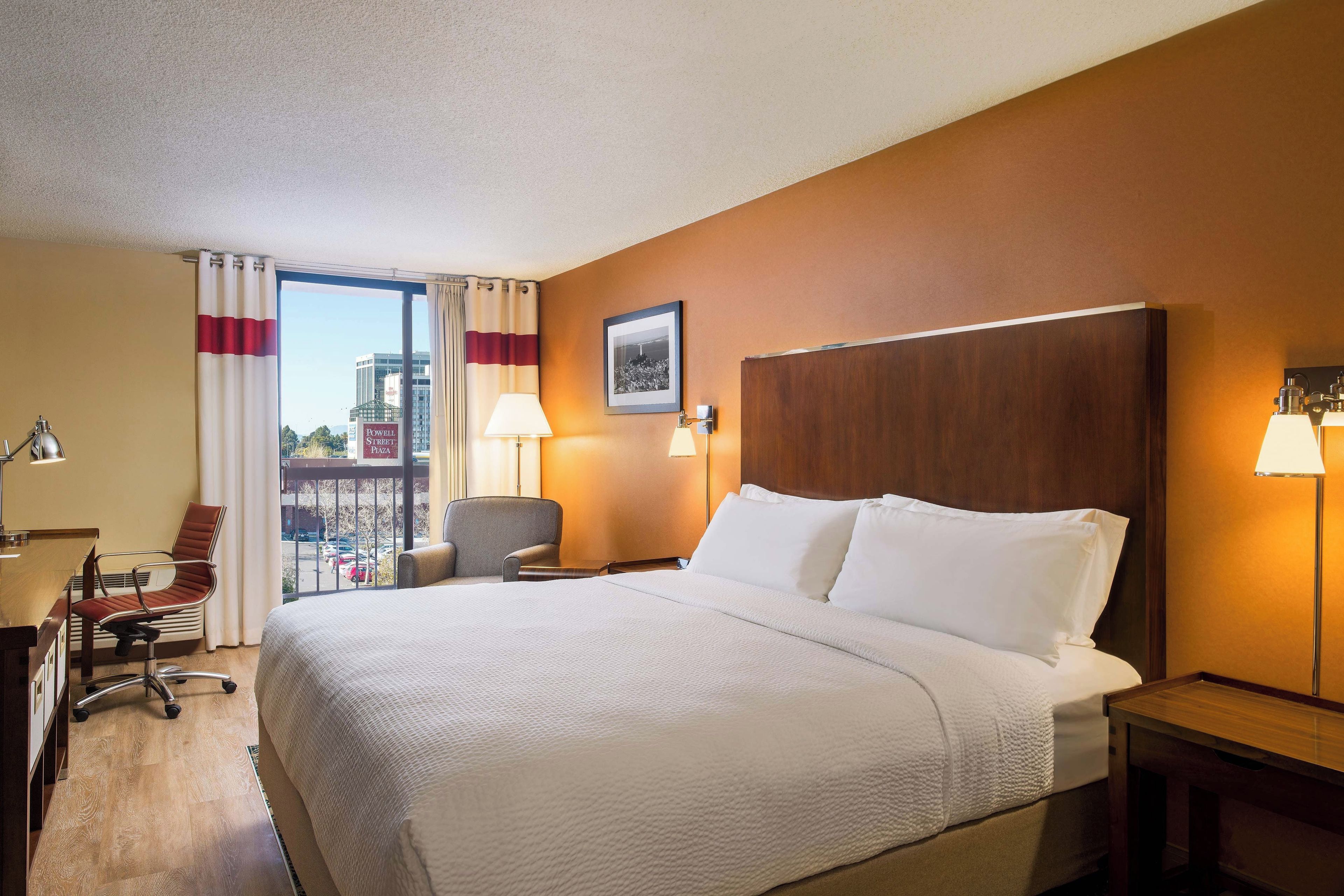 Quarto de hotel com uma cama grande, uma escrivaninha com cadeira, uma luminária de mesa, uma mesa de cabeceira com abajur e vista para o exterior.