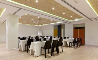 Sala de conferência com várias mesas e cadeiras, equipada com garrafas de água e copos, e um quadro branco na parede.