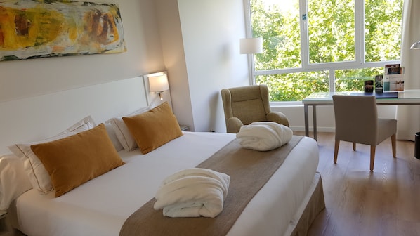 Quarto de hotel com uma cama grande, duas cadeiras, uma escrivaninha e uma janela com vista para árvores.