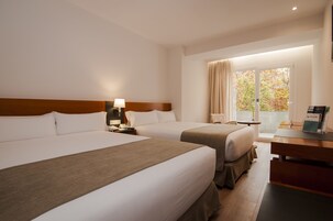 Quarto de hotel com duas camas, uma escrivaninha, uma luminária e uma janela com cortinas.