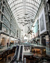 Um centro comercial com vários níveis, balaústres de vidro, escadas rolantes e um teto alto com esculturas de pássaros penduradas.