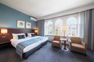 Quarto de hotel com uma cama grande, duas poltronas, uma mesinha e uma janela com cortinas.